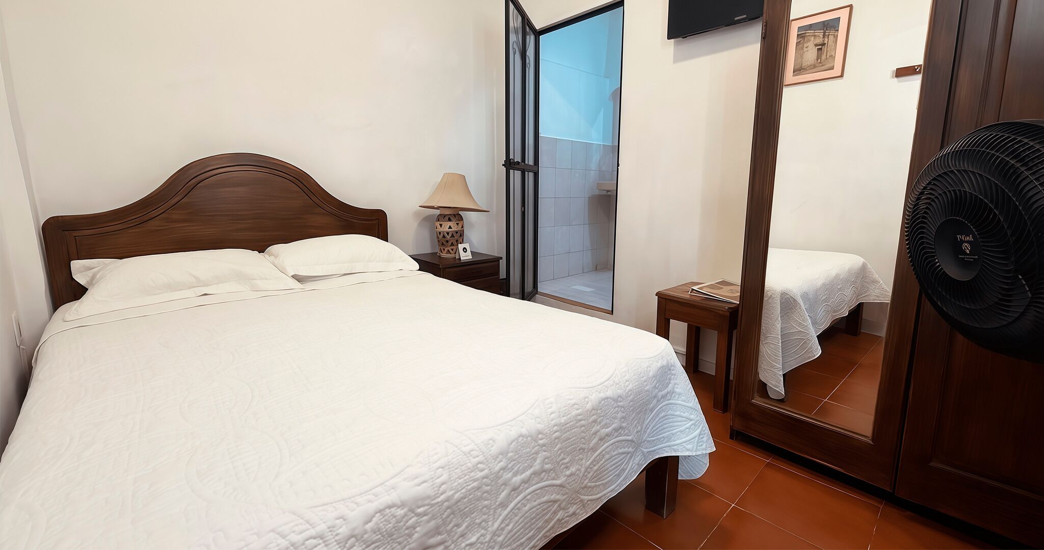 Quarto com cama de madeira, mesa de cabeceira com luminária, um banquinho e um espelho.