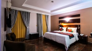 Quarto de hotel com uma cama grande, mesa de cabeceira, uma cadeira e janela com cortinas.