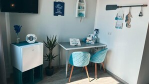 Um espaço de estar compacto com televisão fixada na parede, uma mesinha com duas cadeiras, uma planta em vaso e uma estante com objetos decorativos.