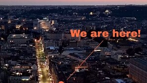 Vista aérea de um horizonte urbano ao entardecer, com uma fonte circular em destaque no primeiro plano, cercada por edifícios iluminados e uma rua movimentada.