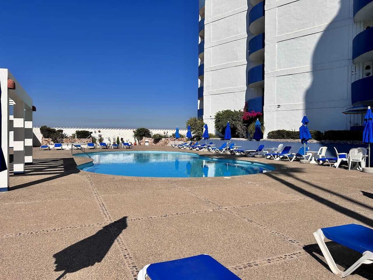 Uma área de piscina com espreguiçadeiras e guarda-sóis, um edifício com varandas, e um céu azul e límpido.