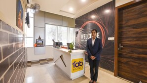 Uma área de recepção de hotel com design moderno, um balcão de recepção com a identificação "Tab Hotels" e uma placa com a palavra "Bem-vindo".