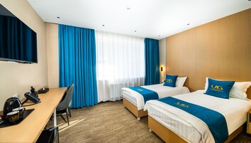 Quarto de hotel com duas camas, uma escrivaninha, uma cadeira, televisão e janela com cortinas.