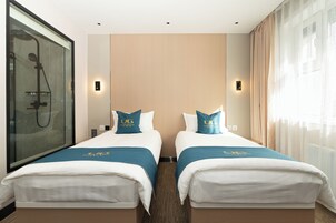 Quarto de hotel com duas camas, área de chuveiro e uma janela grande com cortinas.