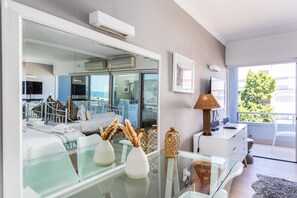 Quarto de hotel moderno com mesa de vidro, mobiliário branco, um espelho grande e vista para o exterior.