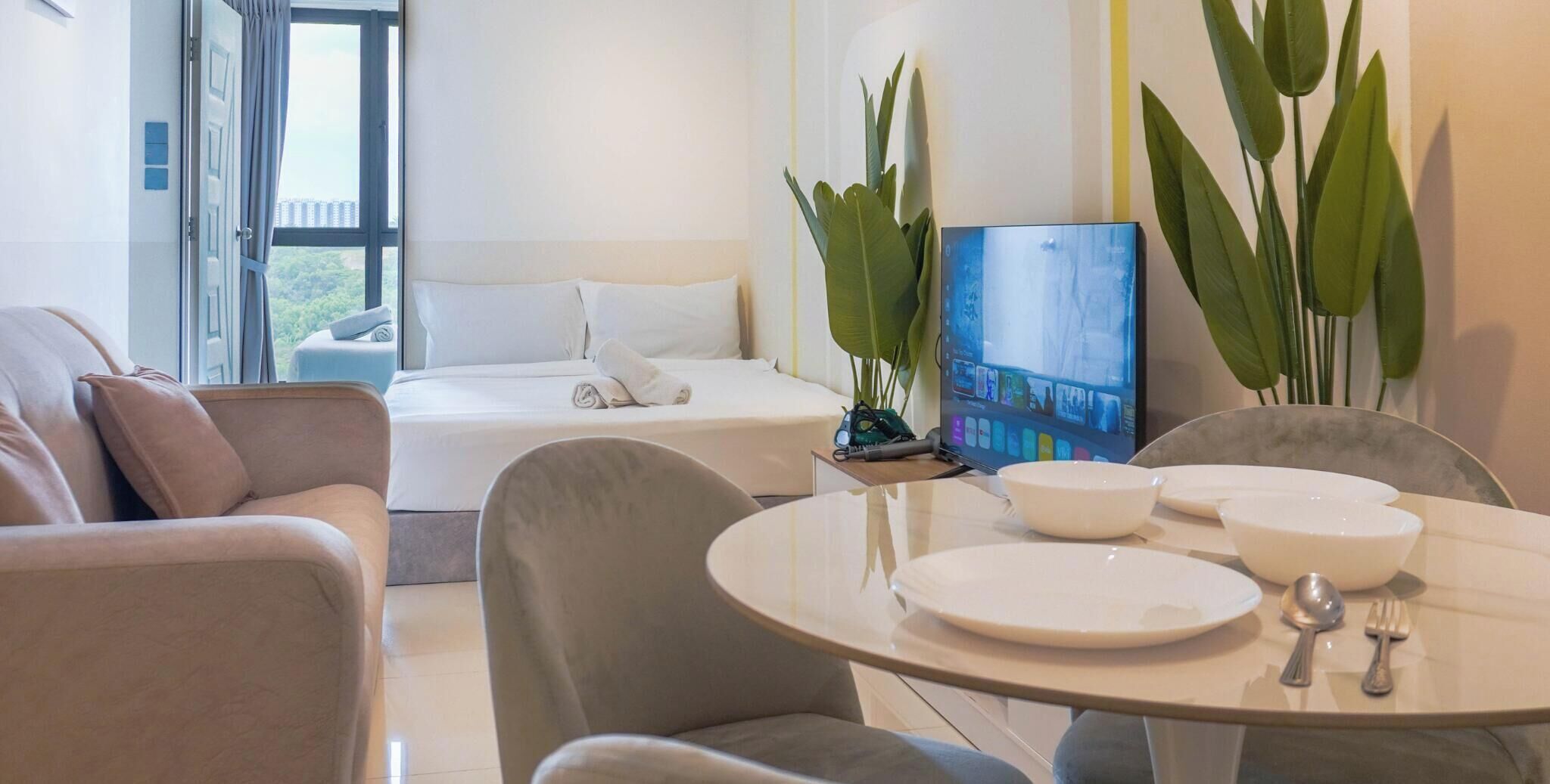 Quarto de hotel moderno com cama, sofá, mesa de jantar com tigelas e talheres, televisão e plantas verdes grandes.