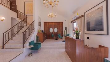 Um hall de hotel com escadaria, balcão de recepção, cadeiras cor turquesa, lustre e obras de arte nas paredes.