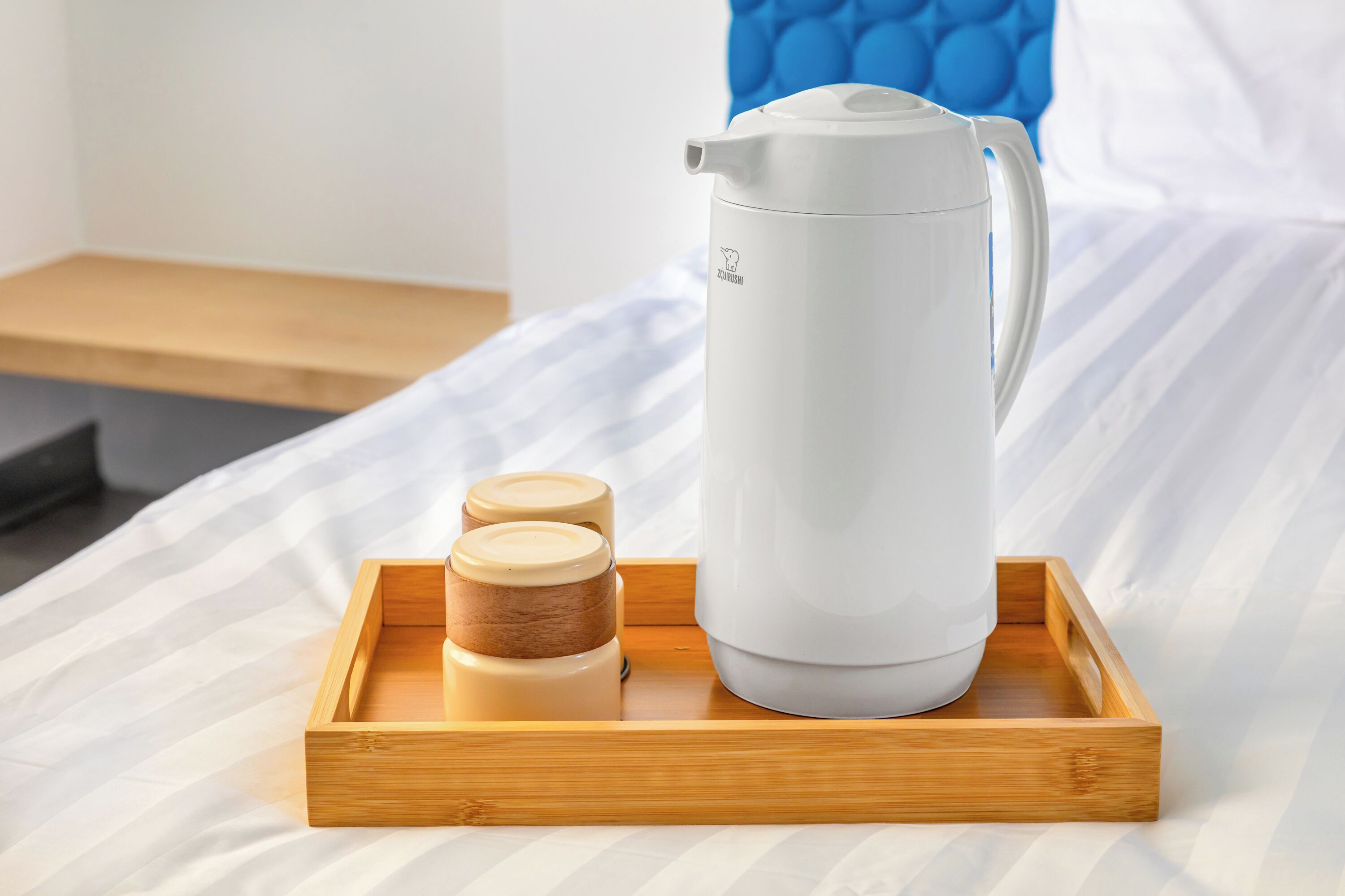 Uma chaleira elétrica branca sobre uma bandeja de madeira em uma cama com roupa de cama listrada.