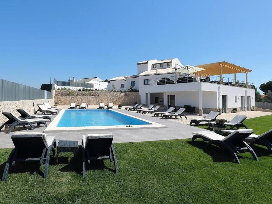 Uma área de piscina moderna ao ar livre, com espreguiçadeiras e um edifício de dois andares ao fundo.
