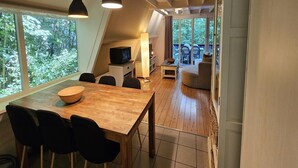 Uma área de refeições com mesa e cadeiras de madeira, uma televisão em um móvel e uma janela grande com vista para o verde.