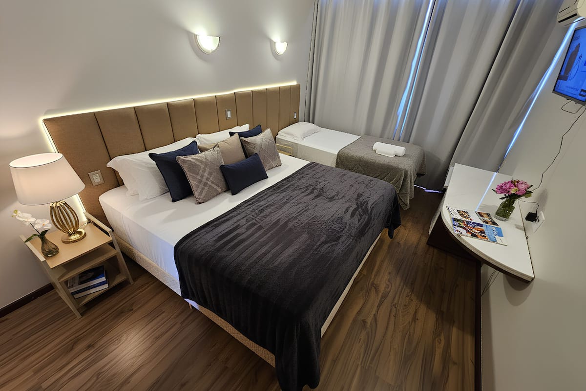 Quarto de hotel com cama, mesas de cabeceira, escrivaninha, abajur e um vaso com flores.