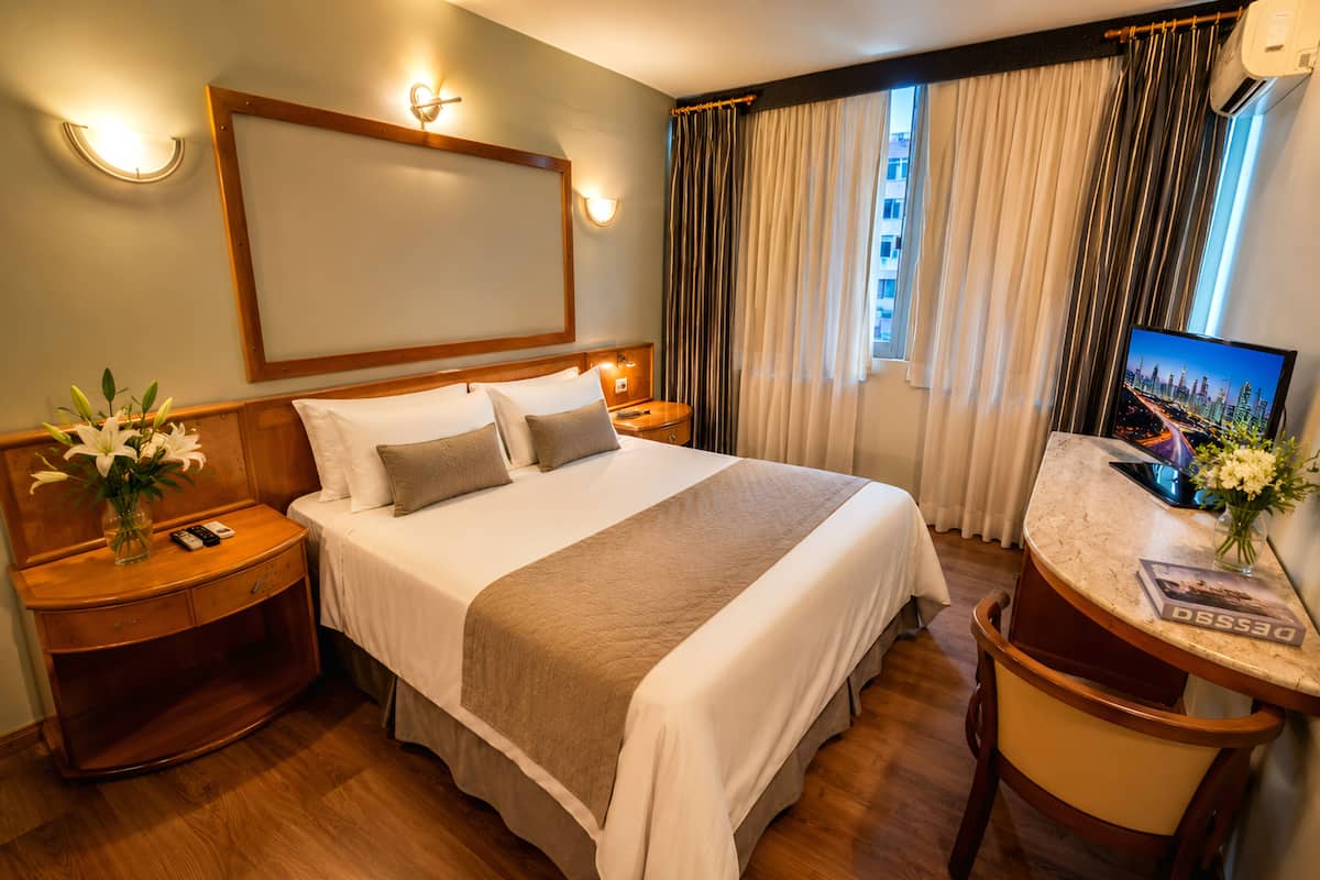 Quarto de hotel com uma cama grande, mesas de cabeceira, uma escrivaninha com cadeira, televisão e uma janela com cortinas.