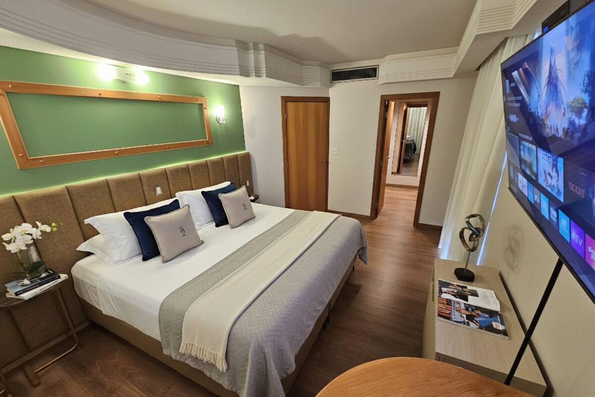 Quarto de hotel com cama, televisão, escrivaninha e espelho.
