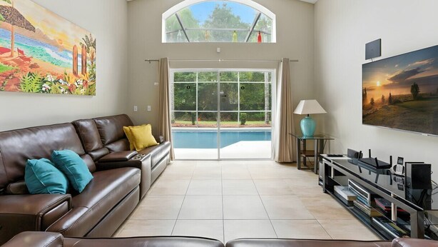 Ampla sala de estar com sofá de couro marrom, televisão e vista para a piscina.