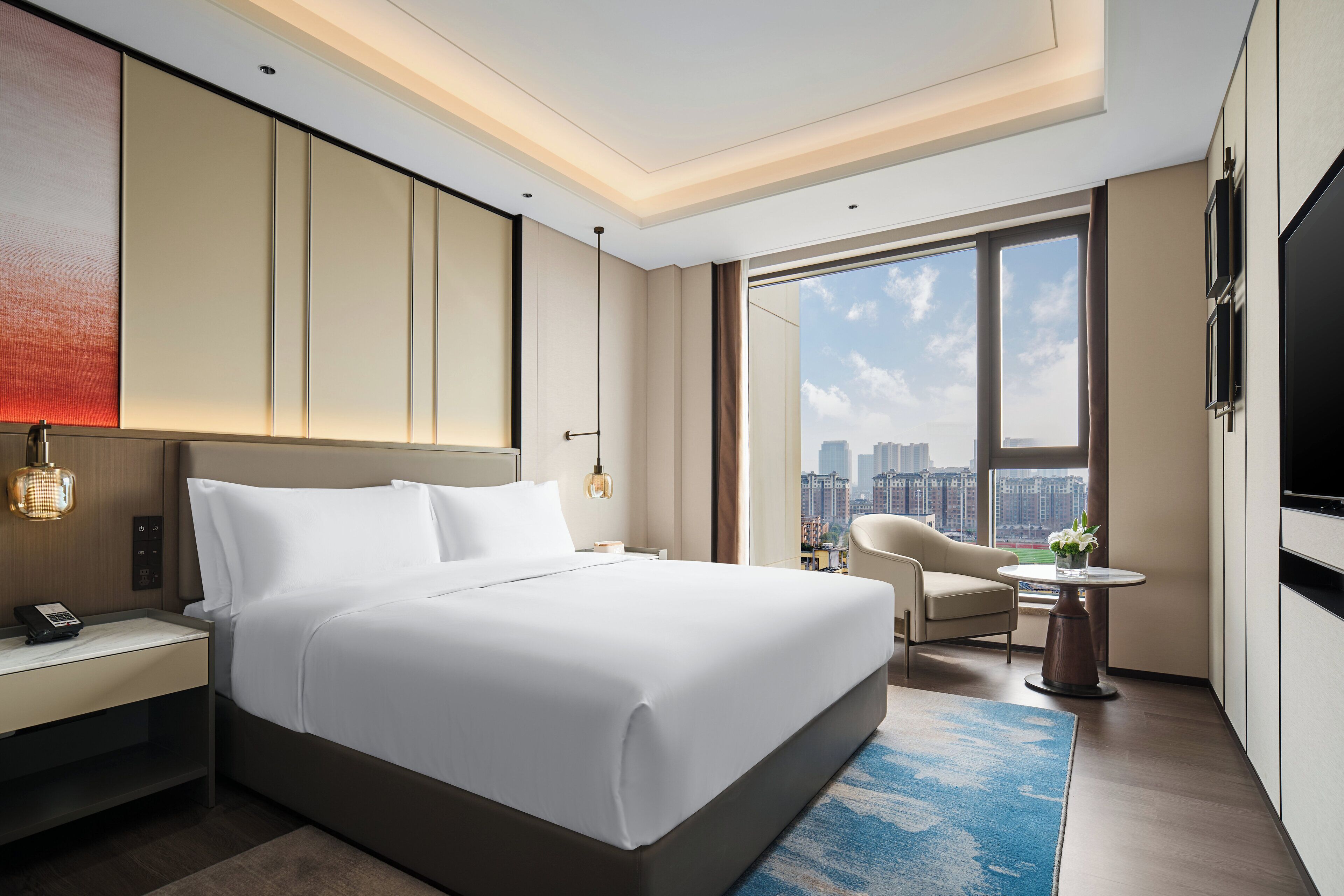 Quarto de hotel moderno com uma cama grande, uma escrivaninha, uma cadeira, uma televisão e vista para a cidade.