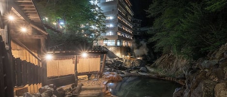 Uma onsen japonesa tradicional, com um balneário de madeira, uma nascente quente de pedra e um edifício de vários andares ao fundo.