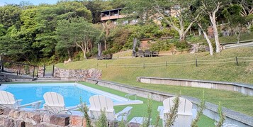 Piscina com espreguiçadeiras, um muro de pedra e uma área gramada com cerca.