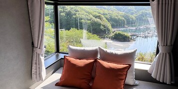 Quarto com uma janela grande que oferece vista para um lago e barcos atracados. Há dois travesseiros em um banco.