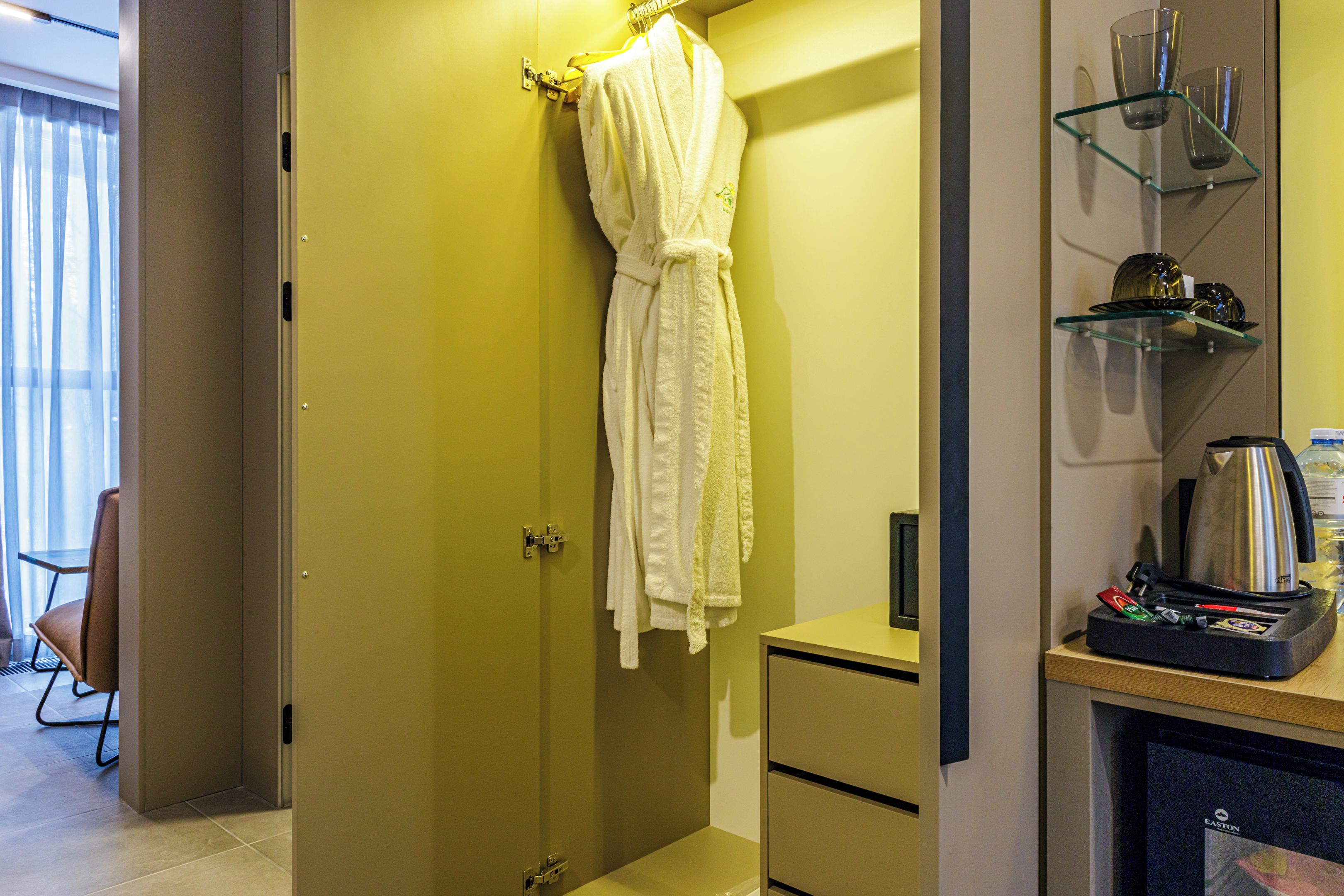 Quarto de hotel com porta amarela, roupão pendurado na porta, minibar e área de estar com uma cadeira.