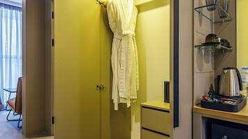 Quarto de hotel com porta amarela, roupão pendurado na porta, minibar e área de estar com uma cadeira.