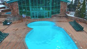 Piscina com formato singular, rodeada por espreguiçadeiras e um edifício de pedra com fachada de vidro.