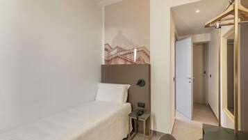 Quarto de hotel com uma cama, uma mesa pequena com telefone, um cabideiro e uma luminária na parede.