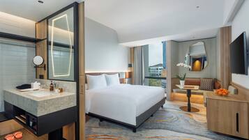 Quarto de hotel moderno com uma cama grande, banheiro com toucador, escrivaninha e vista para a cidade.