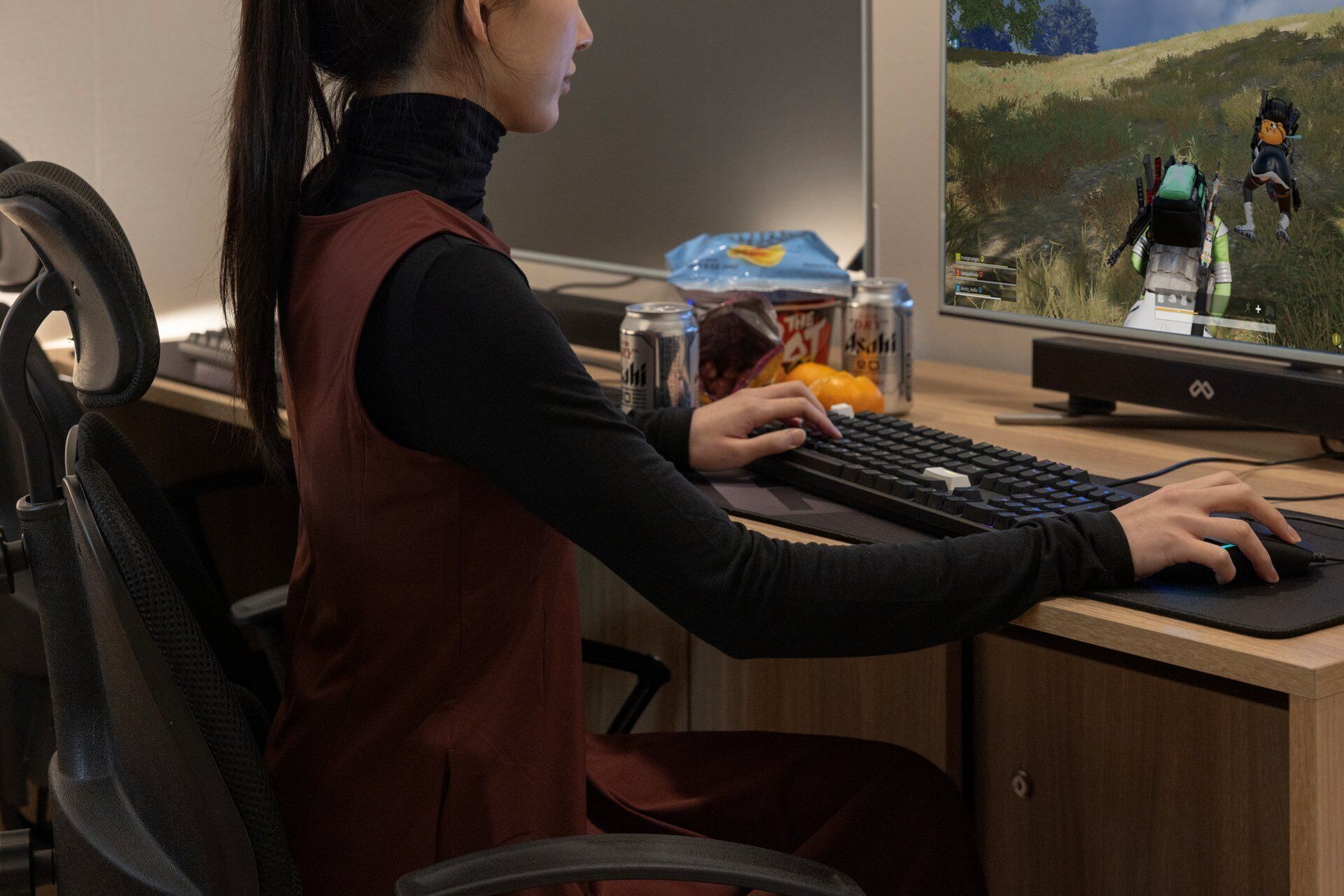 Uma pessoa sentada em uma mesa com um computador, teclado e mouse. Há petiscos e uma lata de refrigerante na mesa.