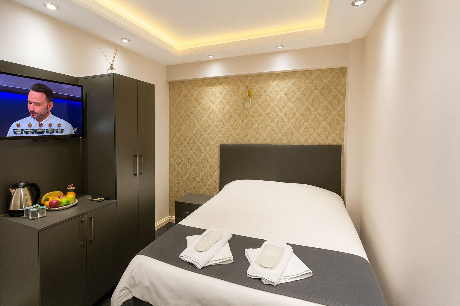 Quarto de hotel com cama, televisão, guarda-roupa e uma pequena mesa com chaleira e frutas.