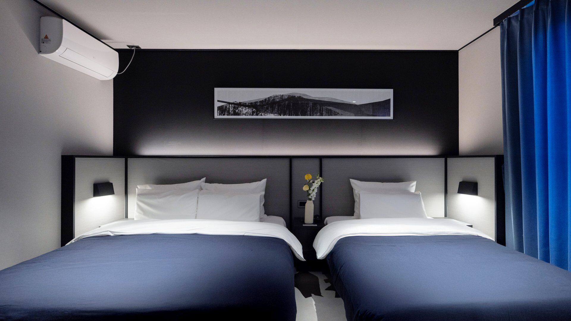Quarto de hotel com duas camas, ar-condicionado fixado na parede, quadro emoldurado e cortina azul.