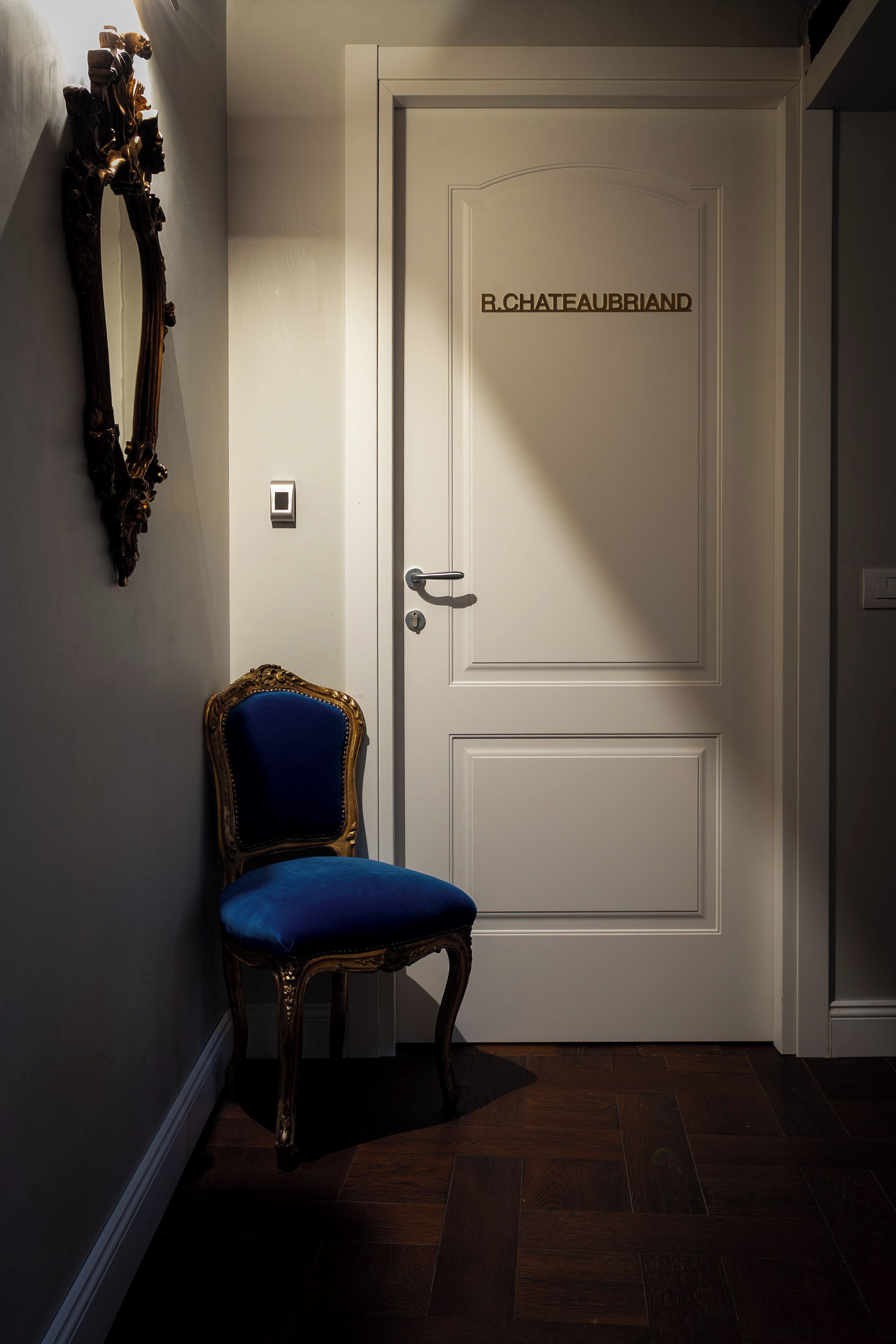 Quarto com porta branca identificada como "B.CHATEAUBRIAND", uma poltrona azul estofada e piso de madeira.