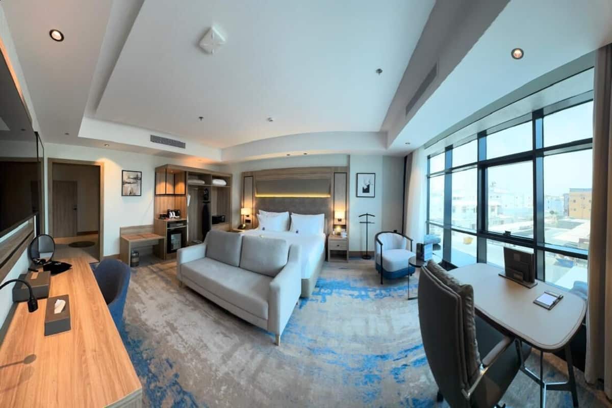 Quarto de hotel moderno com teto curvo, uma cama grande, escrivaninha, cadeira e vista da cidade através de janelas amplas.