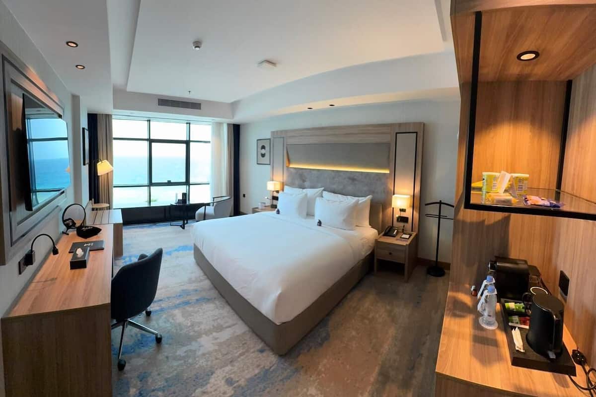 Quarto de hotel moderno com uma cama grande, uma escrivaninha com cadeira, luminária de mesa, cafeteira e vista para o oceano.