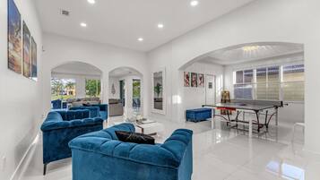 Uma sala de estar ampla com sofás de veludo azul, uma mesa de pingue-pongue e uma grande janela com vista para o exterior.