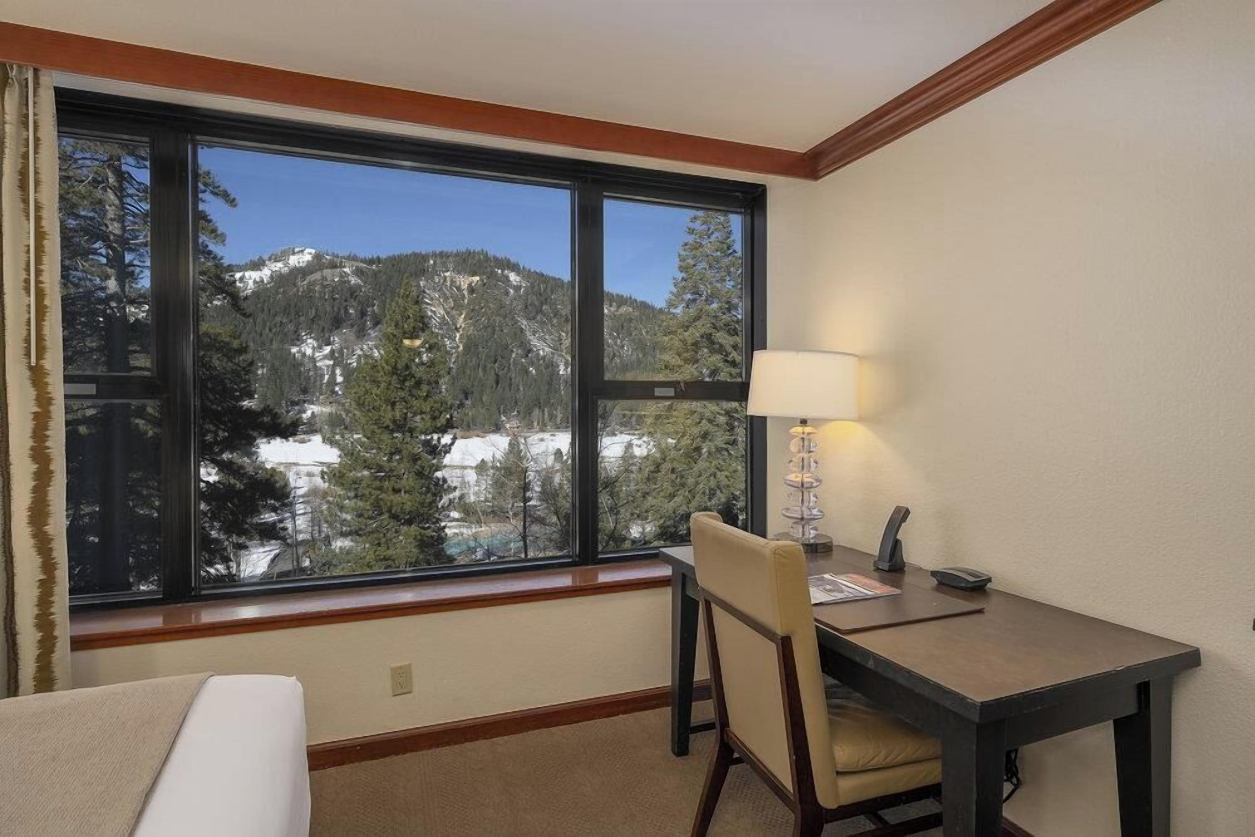 Quarto de hotel com uma grande janela com vista para as montanhas, uma mesa com abajur e uma cadeira.
