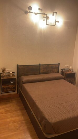Quarto com cabeceira de madeira, cama com colcha marrom, mesa de cabeceira com abajur e luminária na parede.