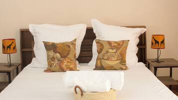 Cama bem arrumada com lençóis brancos, duas almofadas decorativas com estampas de animais e uma cesta de vime com uma toalha.
