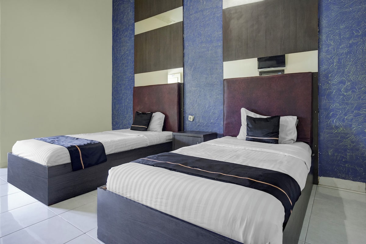Duas camas de solteiro com cabeceiras escuras e roupa de cama branca, posicionadas em frente a uma parede com padrão azul e cinza.