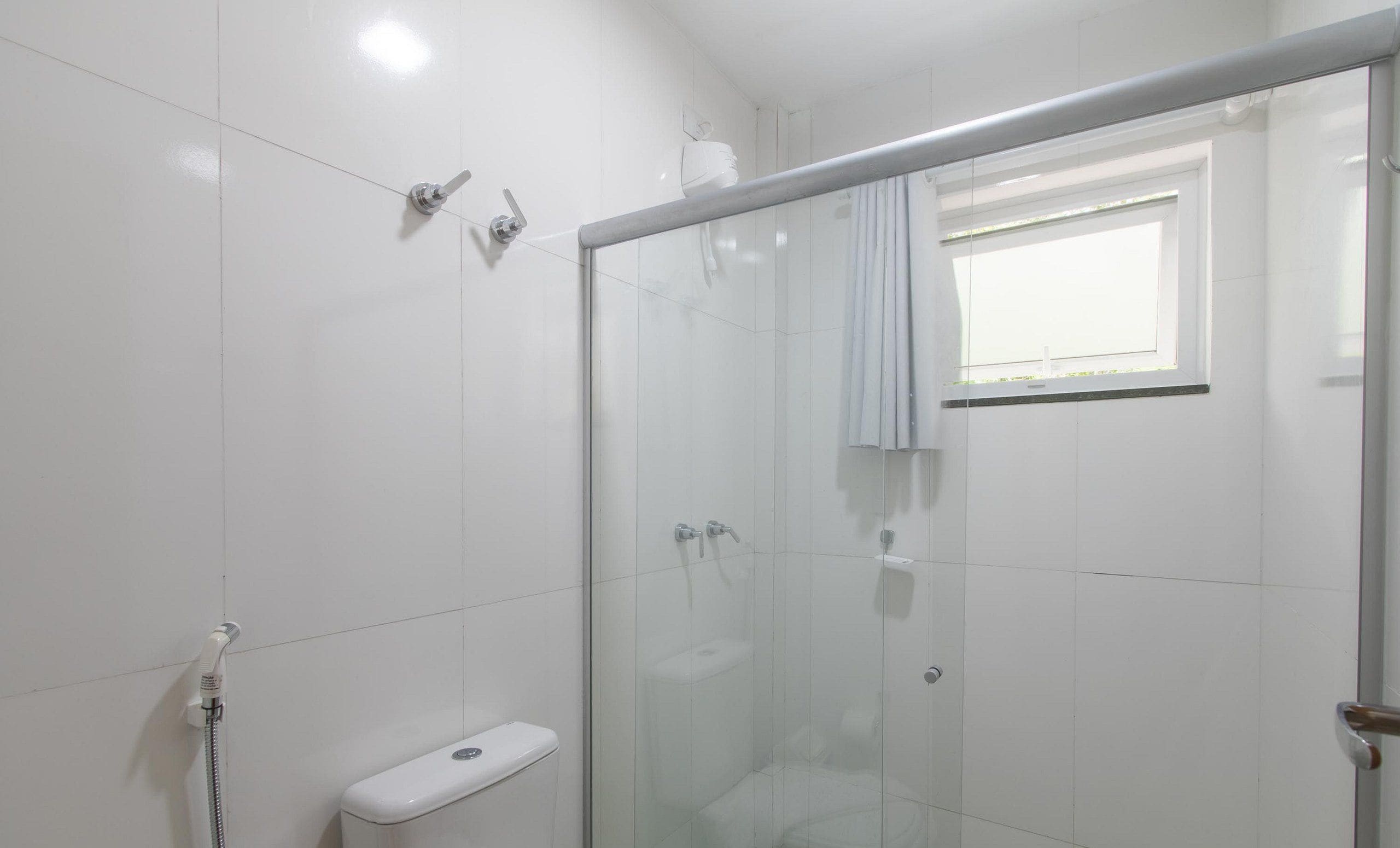 Uma casa de banho moderna com azulejos brancos, cabine de duche em vidro, sanita e janela com cortina.