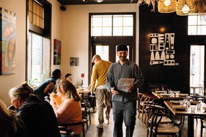 Interior de restaurante com mesas e cadeiras de madeira, janelas amplas e obras de arte nas paredes.