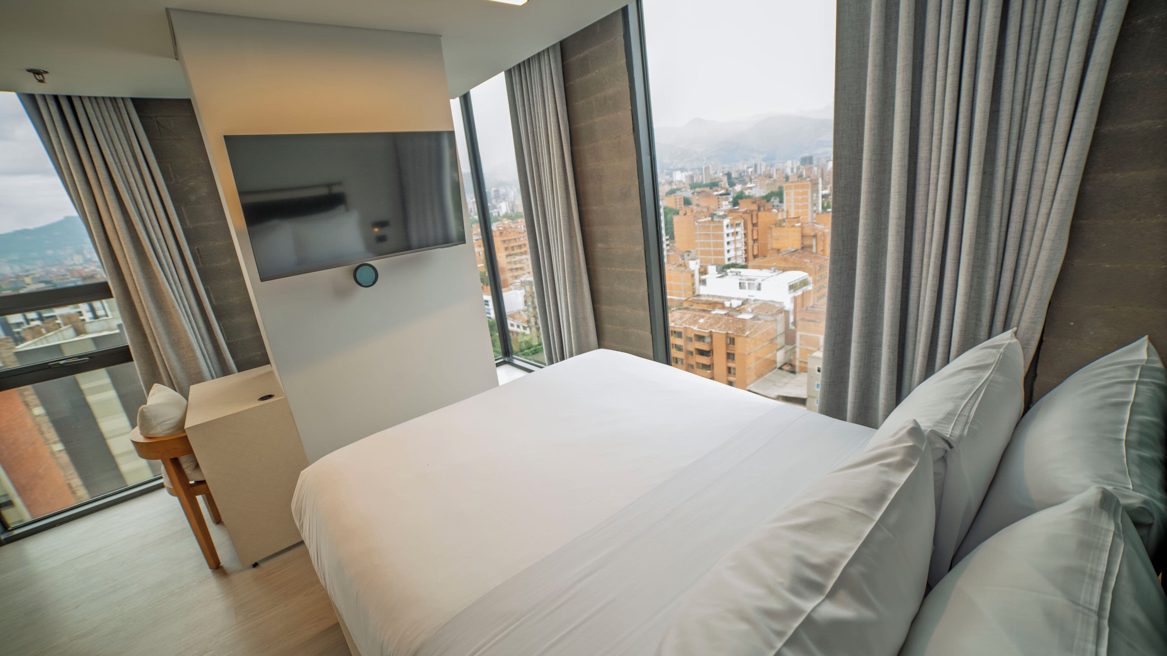 Quarto de hotel com cama, televisão, mesa pequena e vista para a paisagem urbana através de grandes janelas.