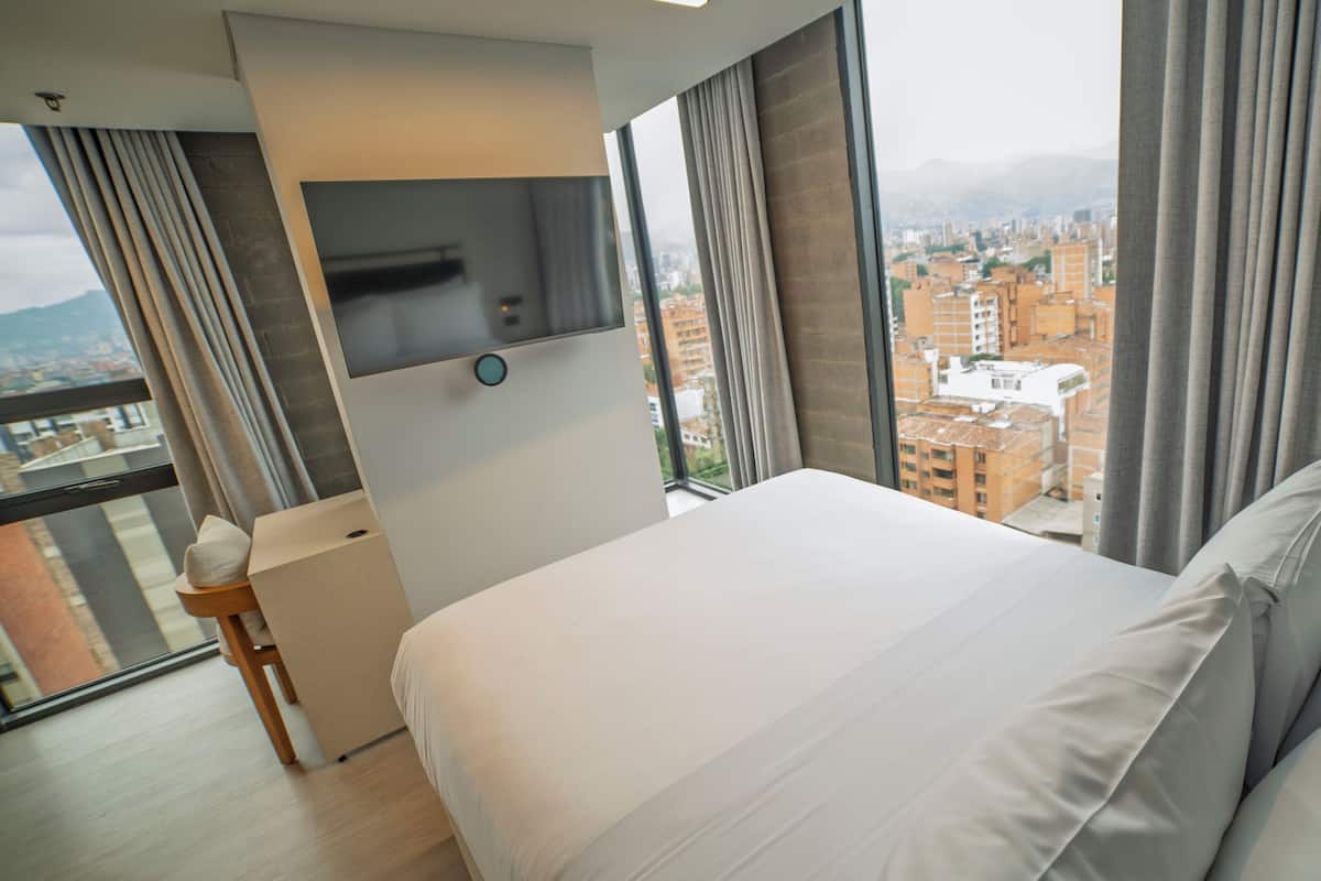 Quarto de hotel com cama, televisão, mesa pequena e vista para a paisagem urbana através de grandes janelas.