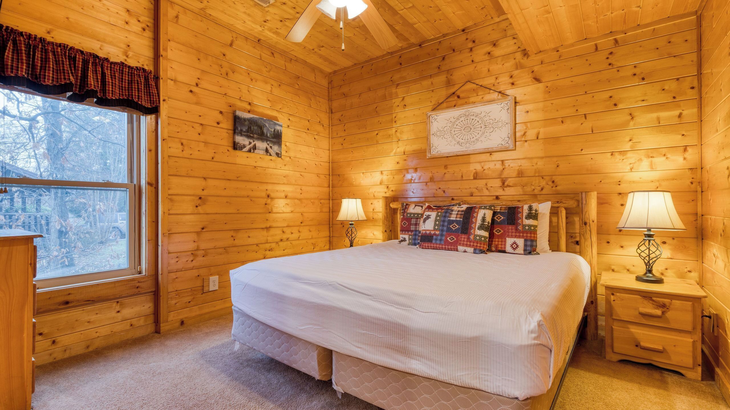 Quarto de cabana de madeira com uma cama, duas mesas de cabeceira, uma janela e um ventilador de teto.