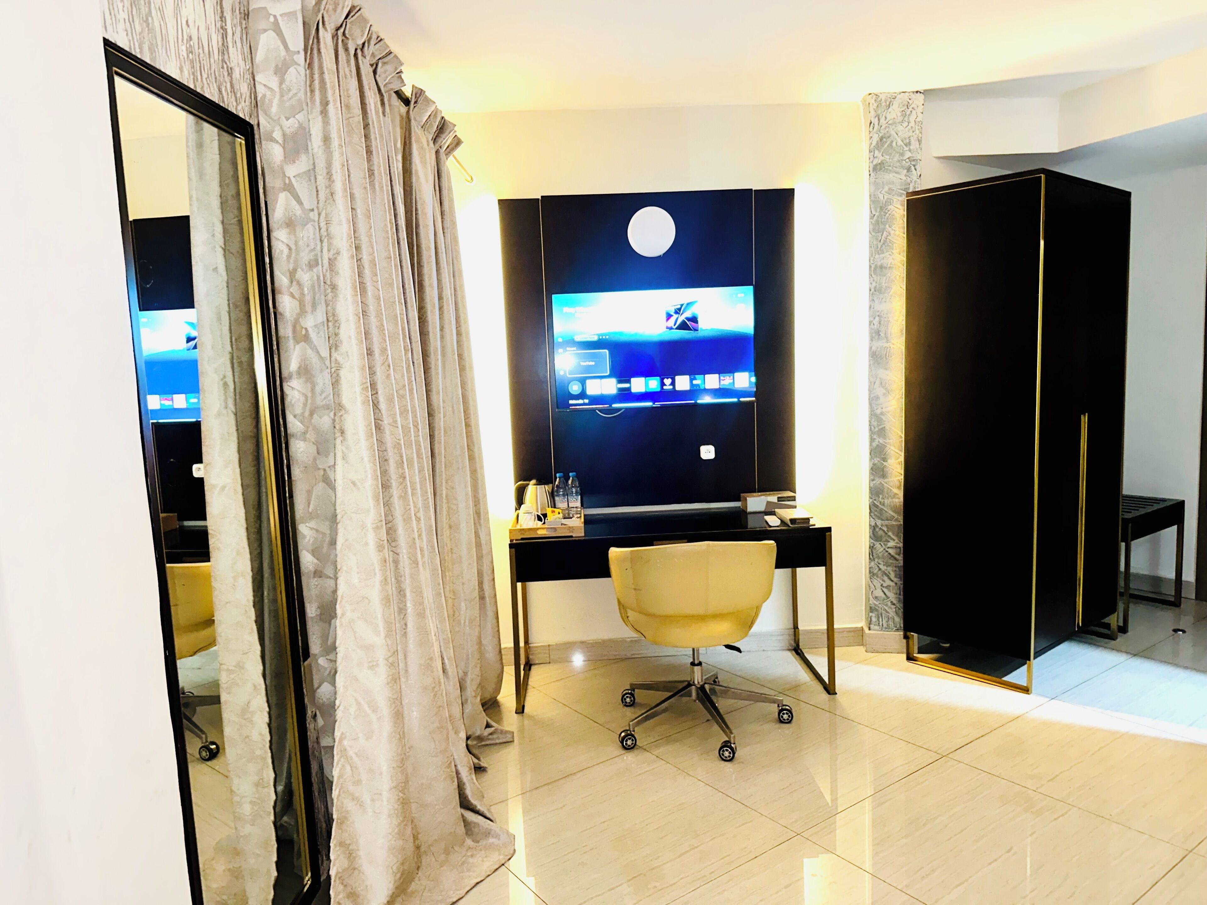 Quarto de hotel com escrivaninha, cadeira, televisão, guarda-roupa e espelho.