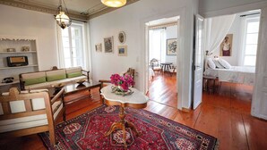 Uma sala de estar ampla com piso de madeira, um tapete vermelho estampado, um sofá branco, uma mesa de centro de madeira com um vaso de flores e um lustre.