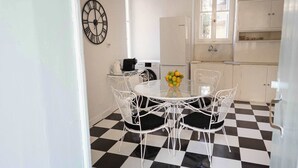 Uma sala de jantar com uma mesa redonda e cadeiras brancas, um relógio na parede, e uma cozinha ao fundo.