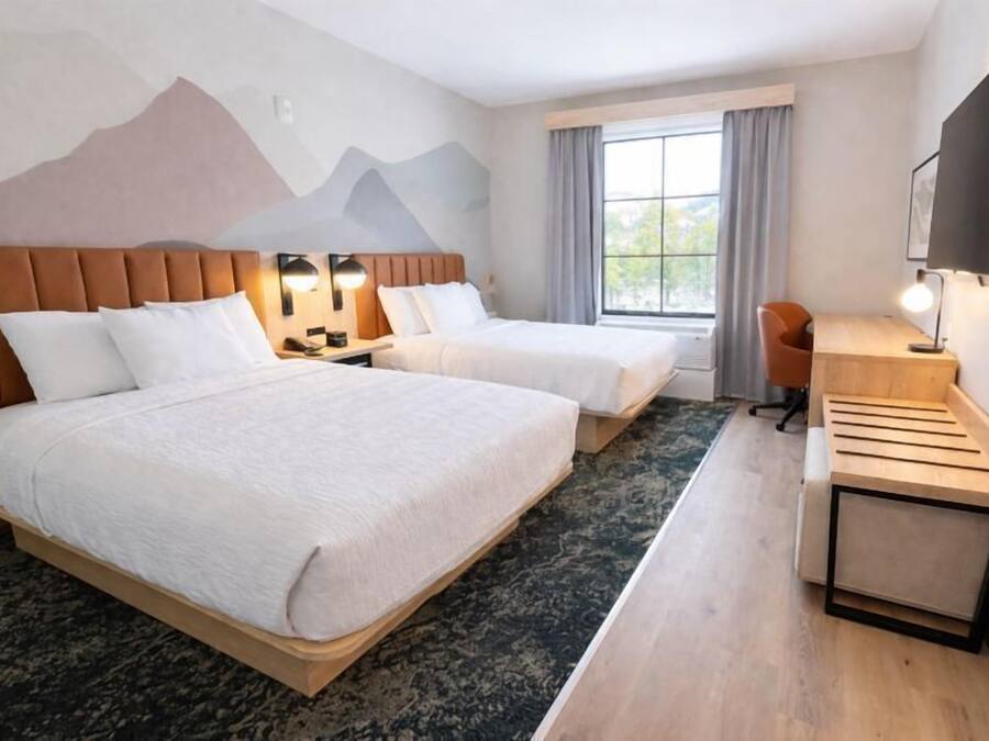 Quarto de hotel com duas camas, uma escrivaninha, uma cadeira, uma televisão e uma janela com cortinas.