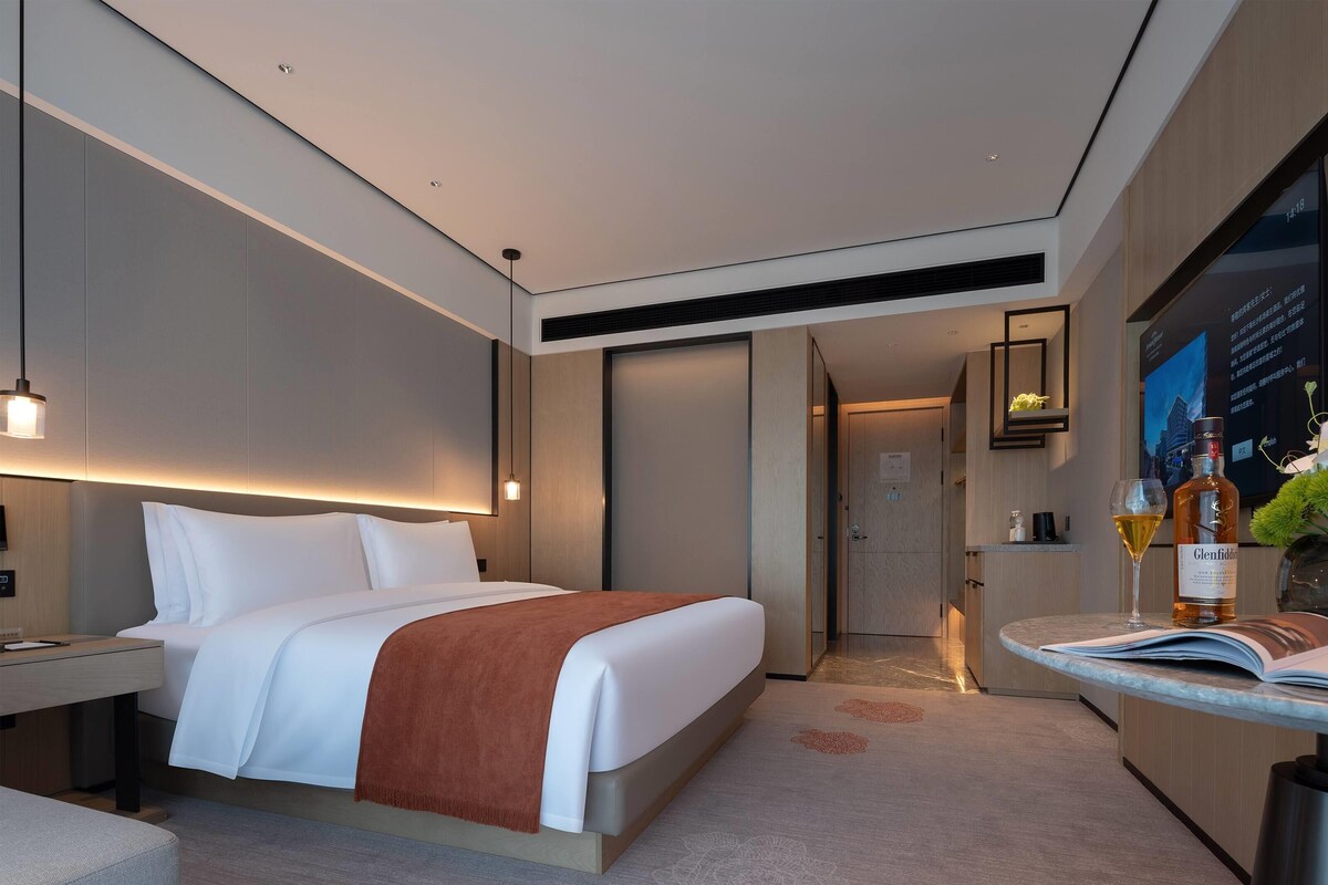 Quarto de hotel moderno com uma cama grande, mesa de cabeceira e um pequeno espaço para sentar. Possui televisão integrada e um minibar.