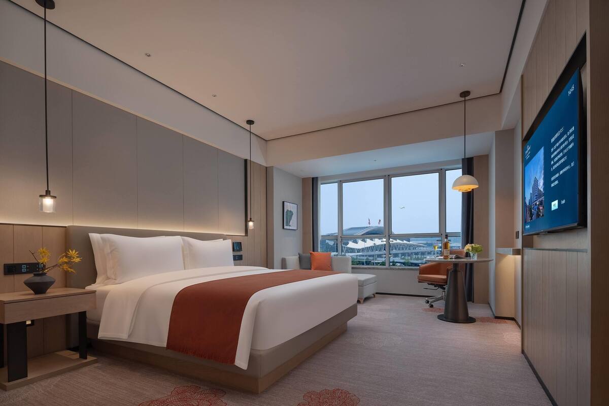 Quarto de hotel moderno com uma cama grande, uma escrivaninha, uma cadeira, uma televisão e uma janela ampla com vista para a cidade.