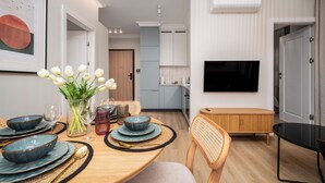 Uma sala de jantar moderna com uma mesa de madeira posta para quatro pessoas, um vaso de flores brancas e uma cozinha ao fundo.